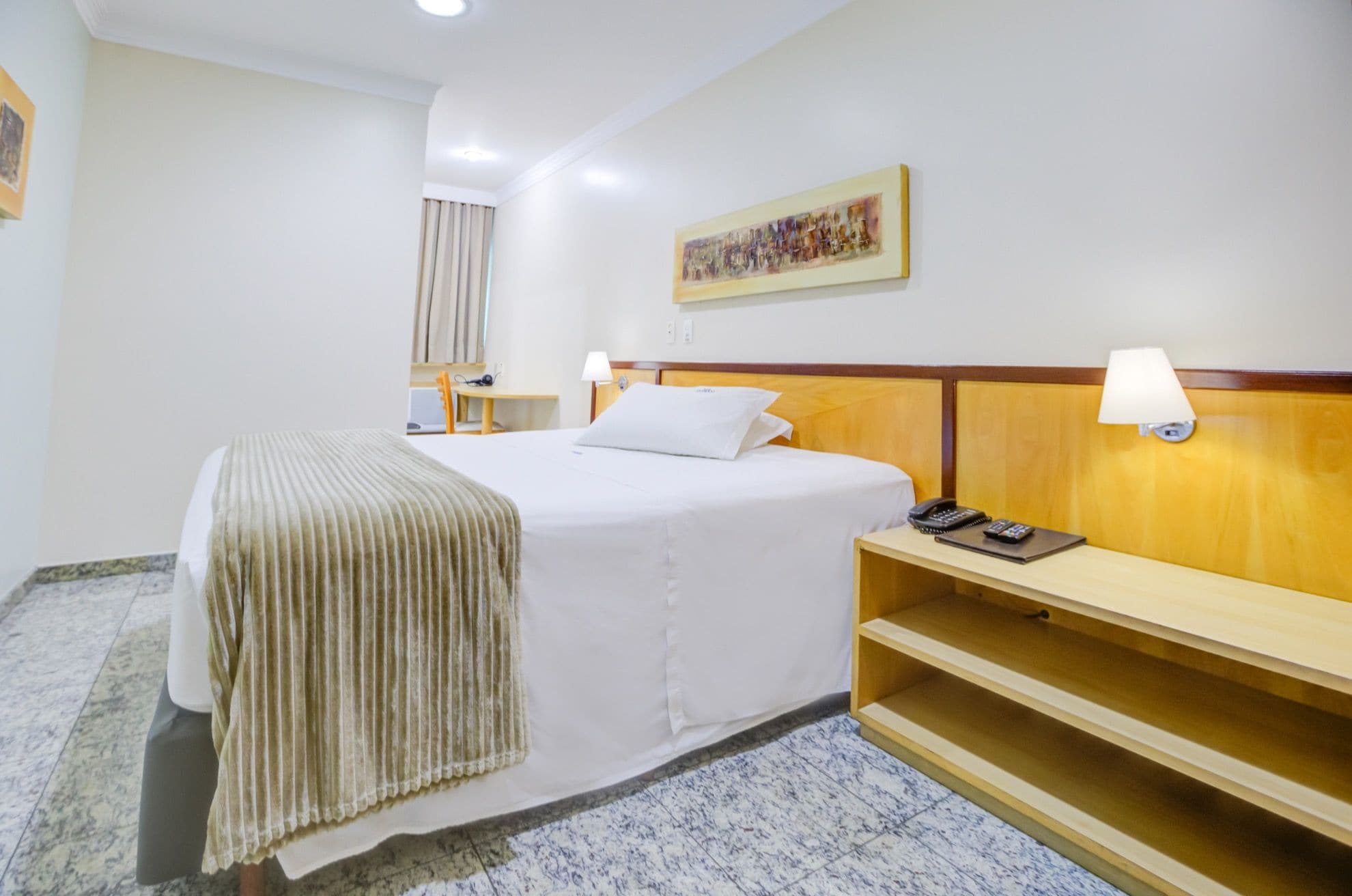 Quarto de hotel com cama, mesa de cabeceira, luminária e um quadro na parede.