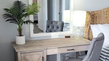 Um quarto com escrivaninha de madeira, cadeira, espelho grande, planta em vaso, cama com roupa de cama listrada e mesa de cabeceira com abajur.