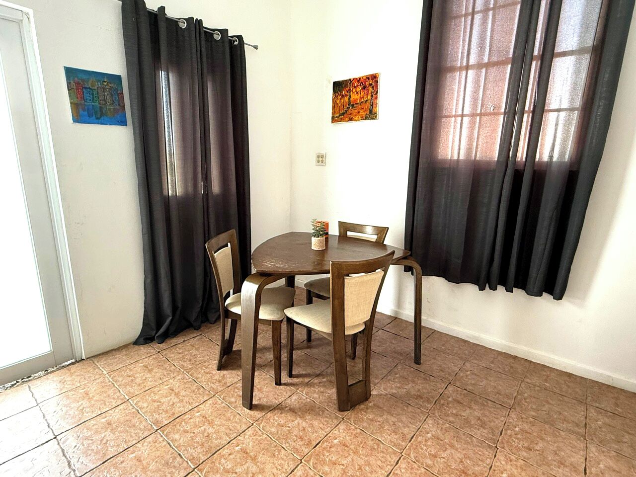 Uma área de refeições com uma mesa redonda e quatro cadeiras, uma porta e duas janelas com cortinas.