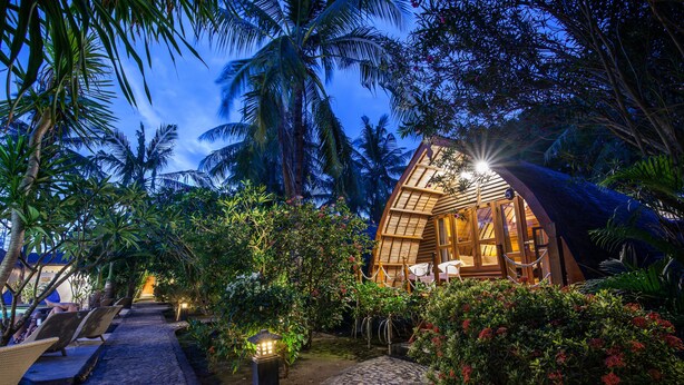 Uma cabana de madeira acolhedora e bem iluminada, cercada por vegetação tropical exuberante e um caminho de pedra que leva a uma área de piscina com espreguiçadeiras.