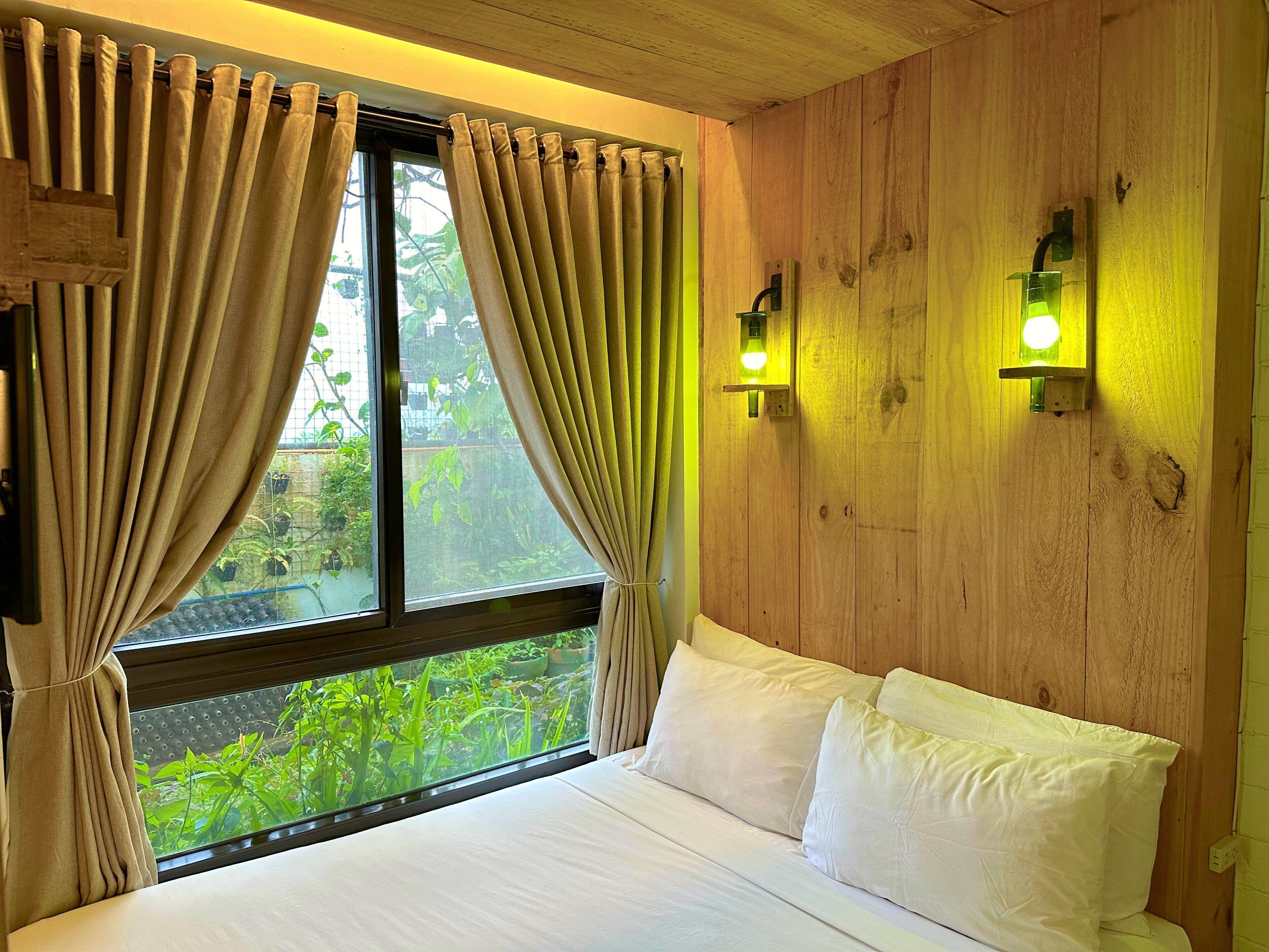 Quarto com parede de madeira, cama com roupa de cama branca e cortinas. Duas luminárias fixadas na parede, de cada lado da janela.