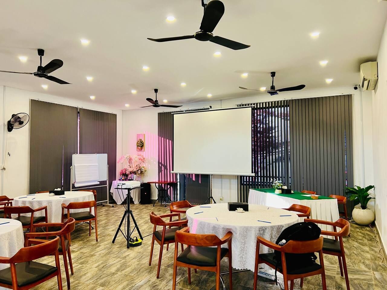 Uma sala de conferências com várias mesas, cadeiras, ventiladores de teto e uma tela grande.