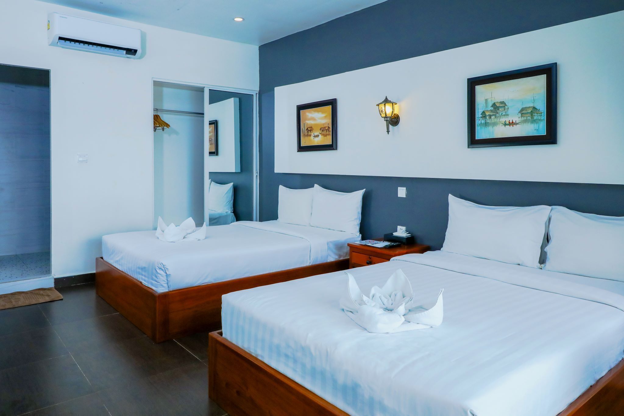 Quarto de hotel com duas camas, cabeceira de madeira, mesa de cabeceira e obras de arte na parede.
