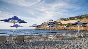 Uma praia com fileiras de espreguiçadeiras e guarda-sóis, um penhasco rochoso ao fundo e o mar em primeiro plano.