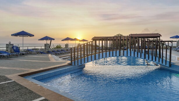 Piscina com deck de madeira e espreguiçadeiras, cercada por guarda-sóis, sob um céu ao pôr do sol.
