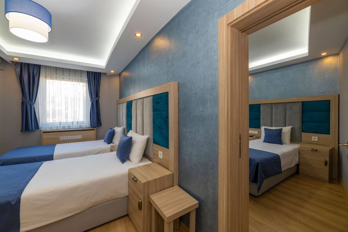 Quarto de hotel com duas camas, uma mesinha de cabeceira, um espelho e uma janela com cortinas.