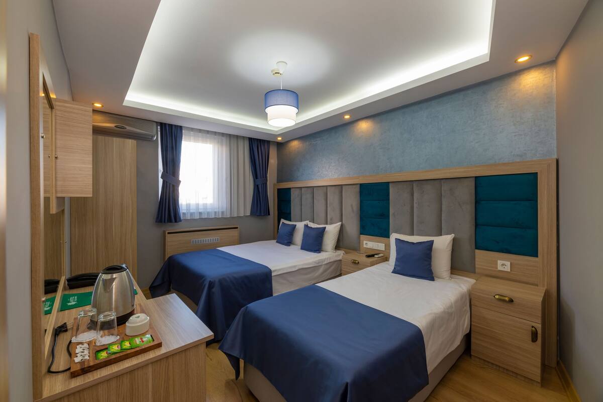 Quarto de hotel com duas camas, uma mesa de trabalho com cafeteira e uma janela com cortinas.