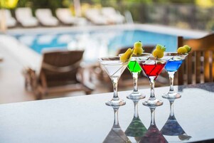 Diversos copos de martini com líquidos coloridos e rodelas de limão sobre uma superfície reflexiva, com uma piscina e espreguiçadeiras ao fundo.