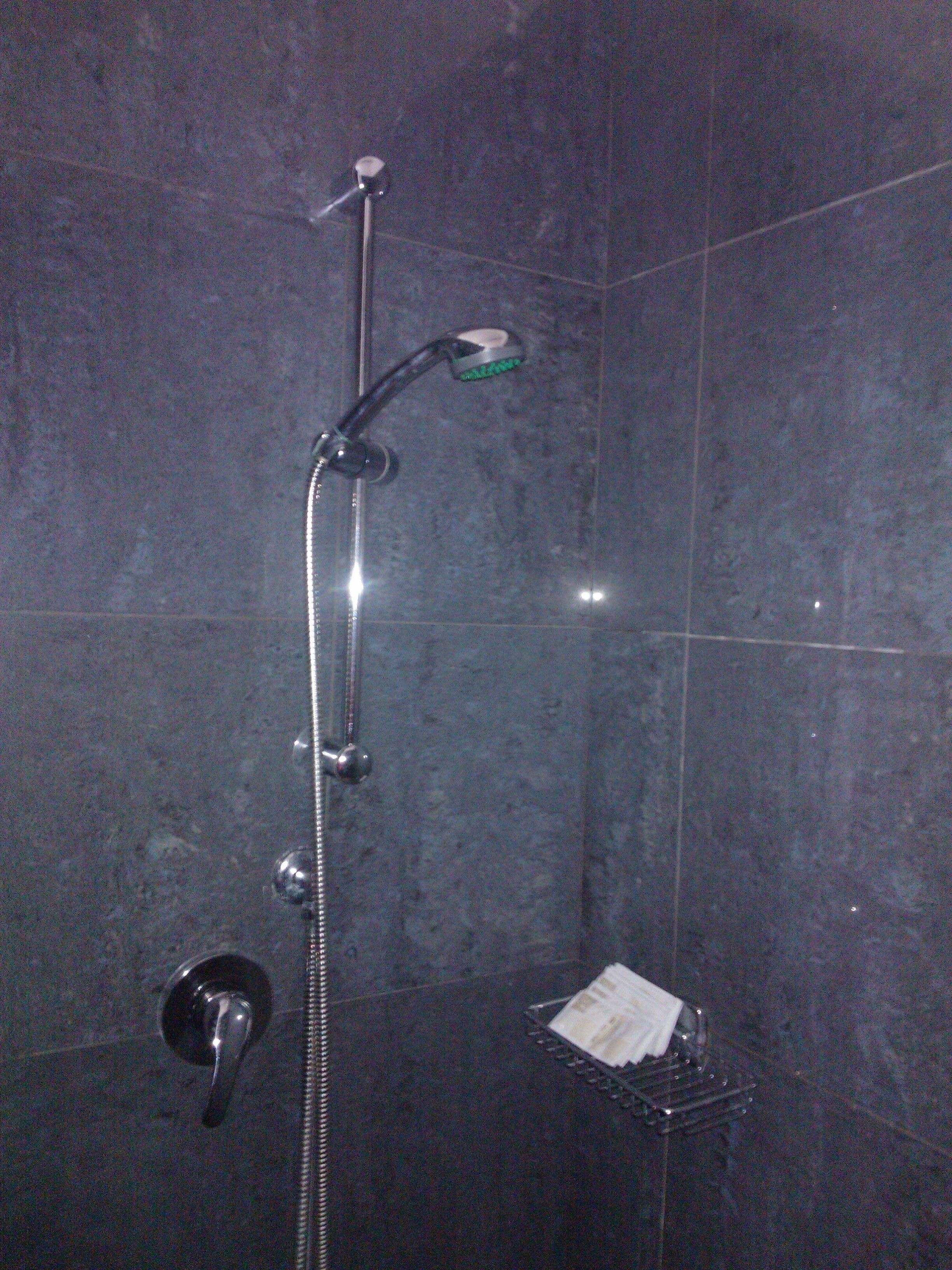 Chuveiro com chuveiro manual, torneira fixada na parede e uma pequena prateleira com lenços.