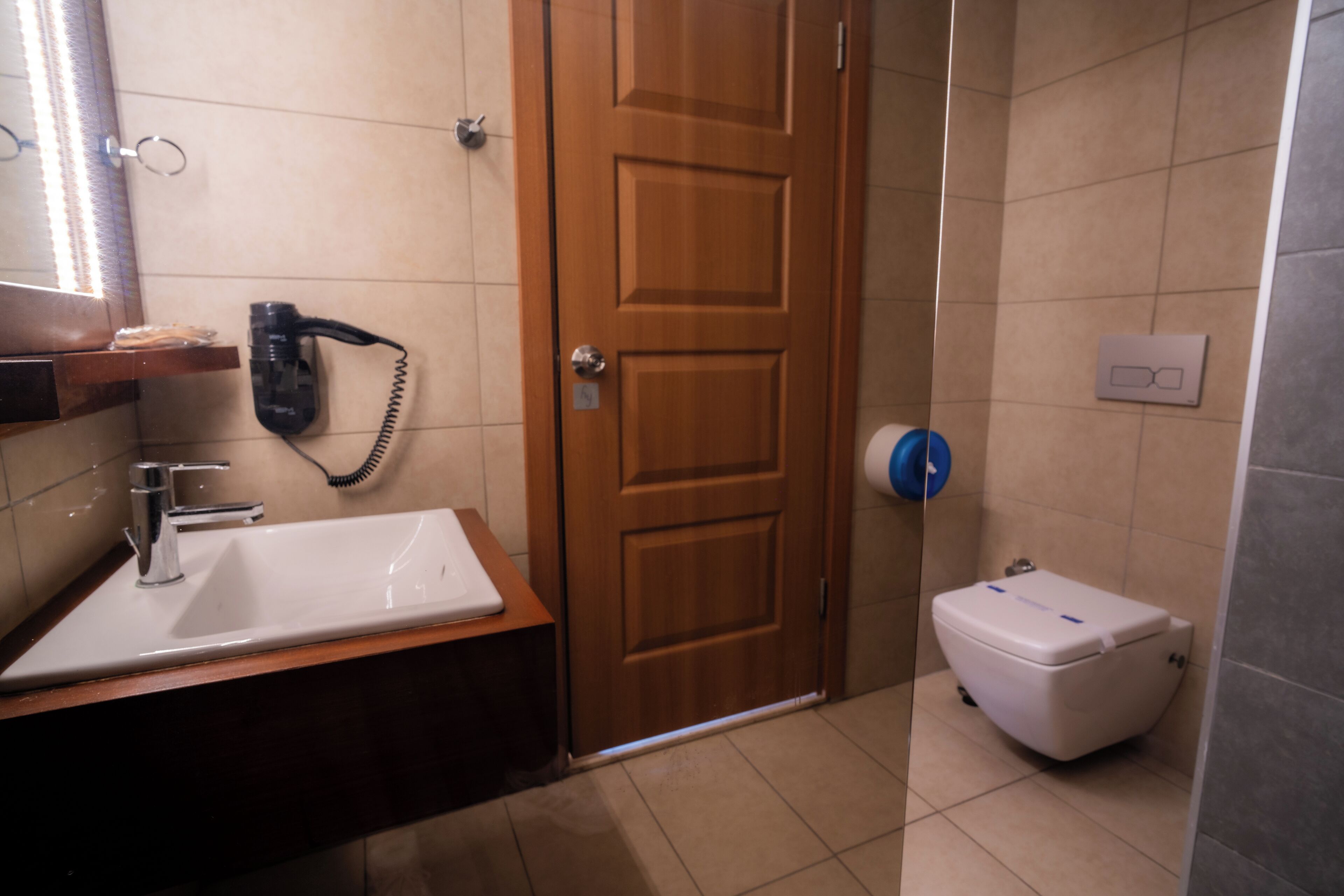 Casa de banho com porta de madeira, lavatório branco, secador de cabelo e sanita.
