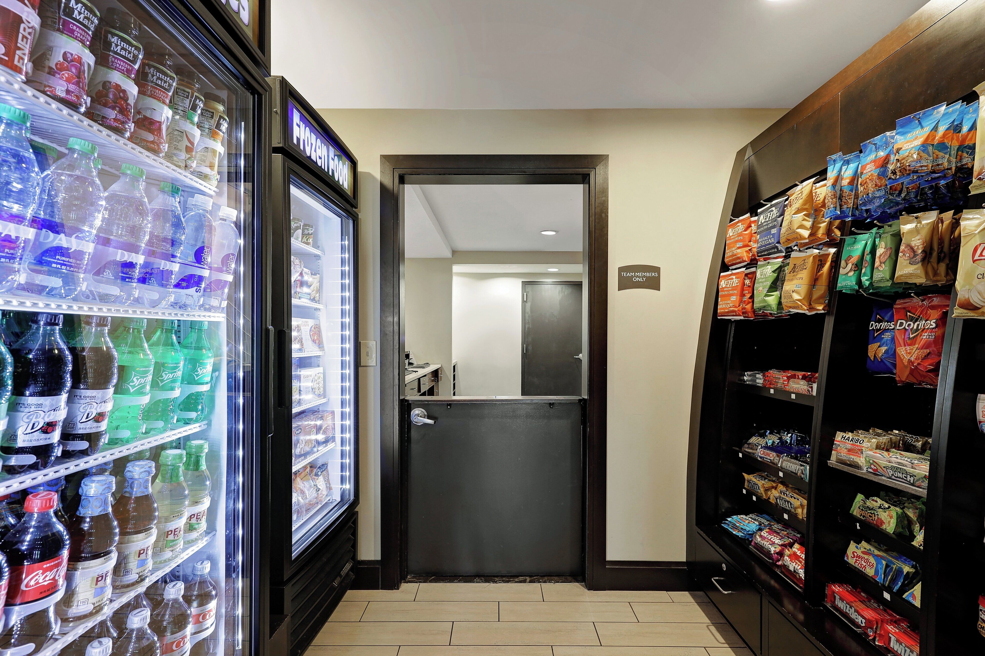 Corredor de hotel com um refrigerador de bebidas repleto de diversas opções, uma máquina de snacks e uma porta que dá para um quarto.