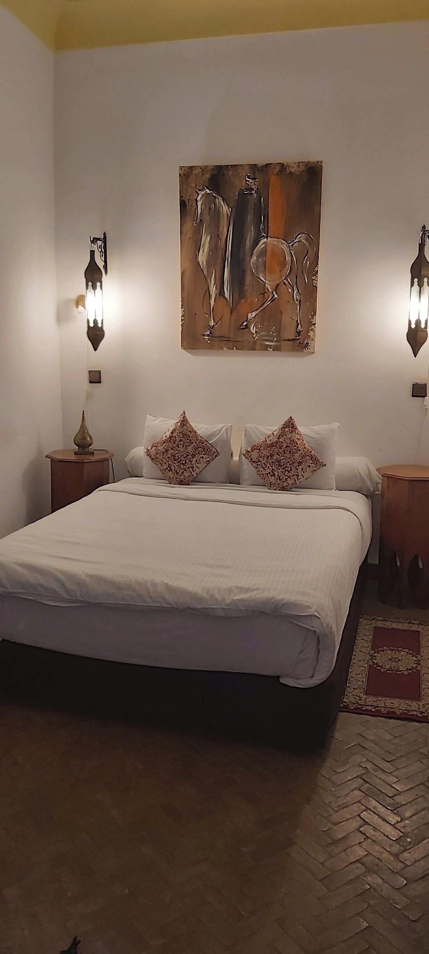Quarto com uma cama, duas luminárias fixadas na parede, um quadro e um tapete estampado.