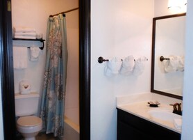 Uma casa de banho com sanita, uma cortina de chuveiro com padrão floral, um porta-toalhas com toalhas, uma pia com espelho e um saboneteira.