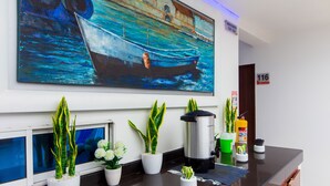 Uma área de recepção de hotel com uma pintura de barco na água, plantas em vasos, uma máquina de café e uma placa indicando o quarto 116.