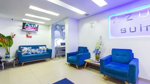 Uma área de recepção moderna com um sofá azul, uma poltrona azul e uma mesa pequena com uma revista. Uma televisão está montada na parede, e há um letreiro que diz "AZU SUIT".