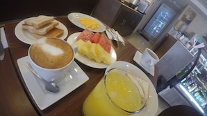 Uma mesa de café da manhã com torradas, ovos mexidos, melancia, abacaxi, um copo de suco de laranja e uma xícara de café.
