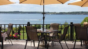 Um terraço com área de estar ao ar livre, incluindo mesas e cadeiras, com vista para um corpo d'água com barcos e uma linha costeira distante.