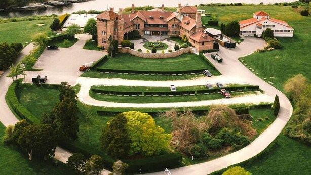 Uma grande mansão, semelhante a um castelo, com um jardim bem cuidado, um caminho circular e um corpo d'água ao fundo.