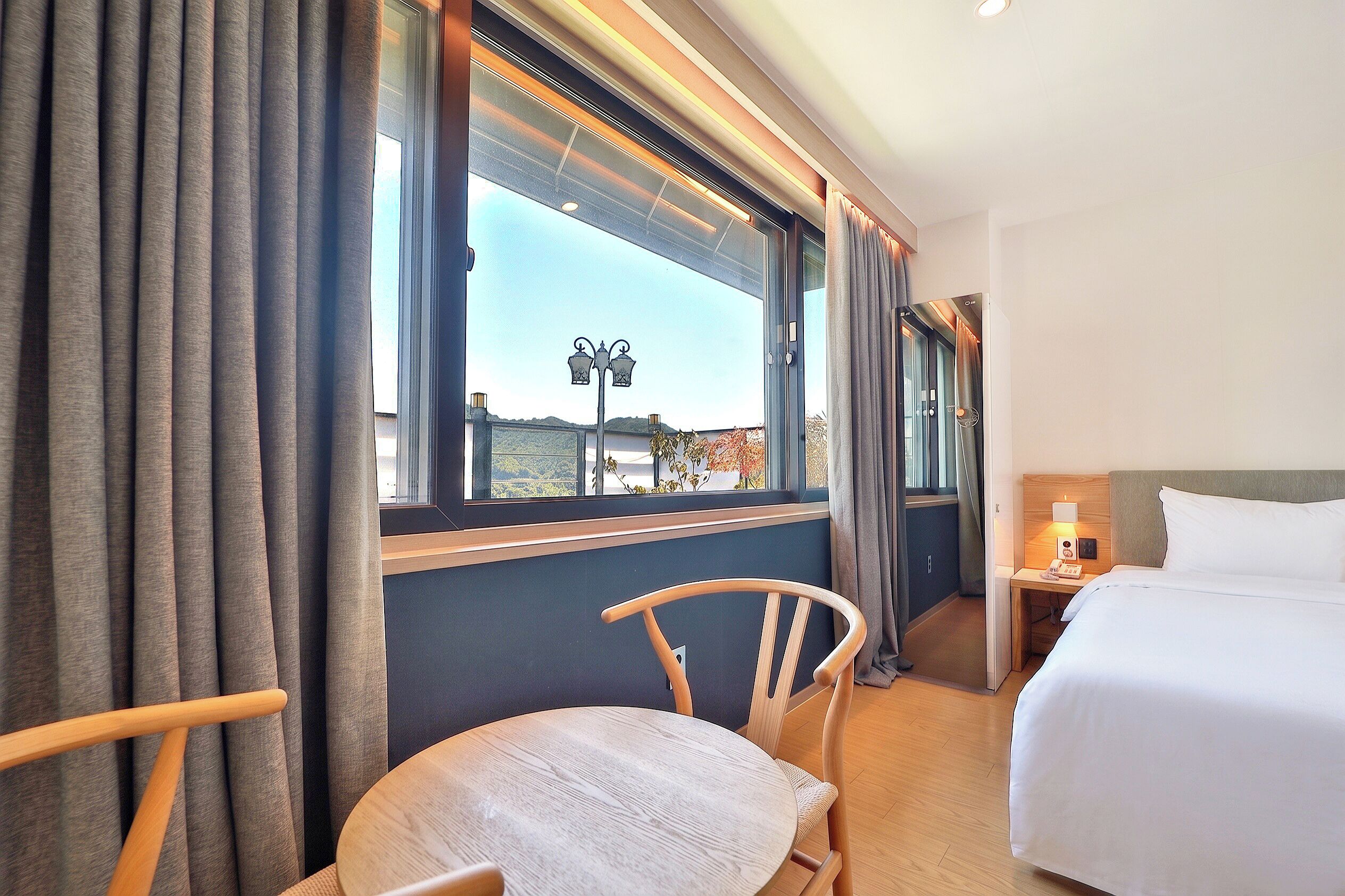 Quarto de hotel com cama, cadeira, escrivaninha com luminária e janela com cortinas.