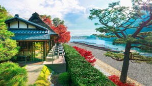 Um edifício tradicional japonês com telhado de palha, área de estar coberta ao ar livre e vista para um lago com barcos.