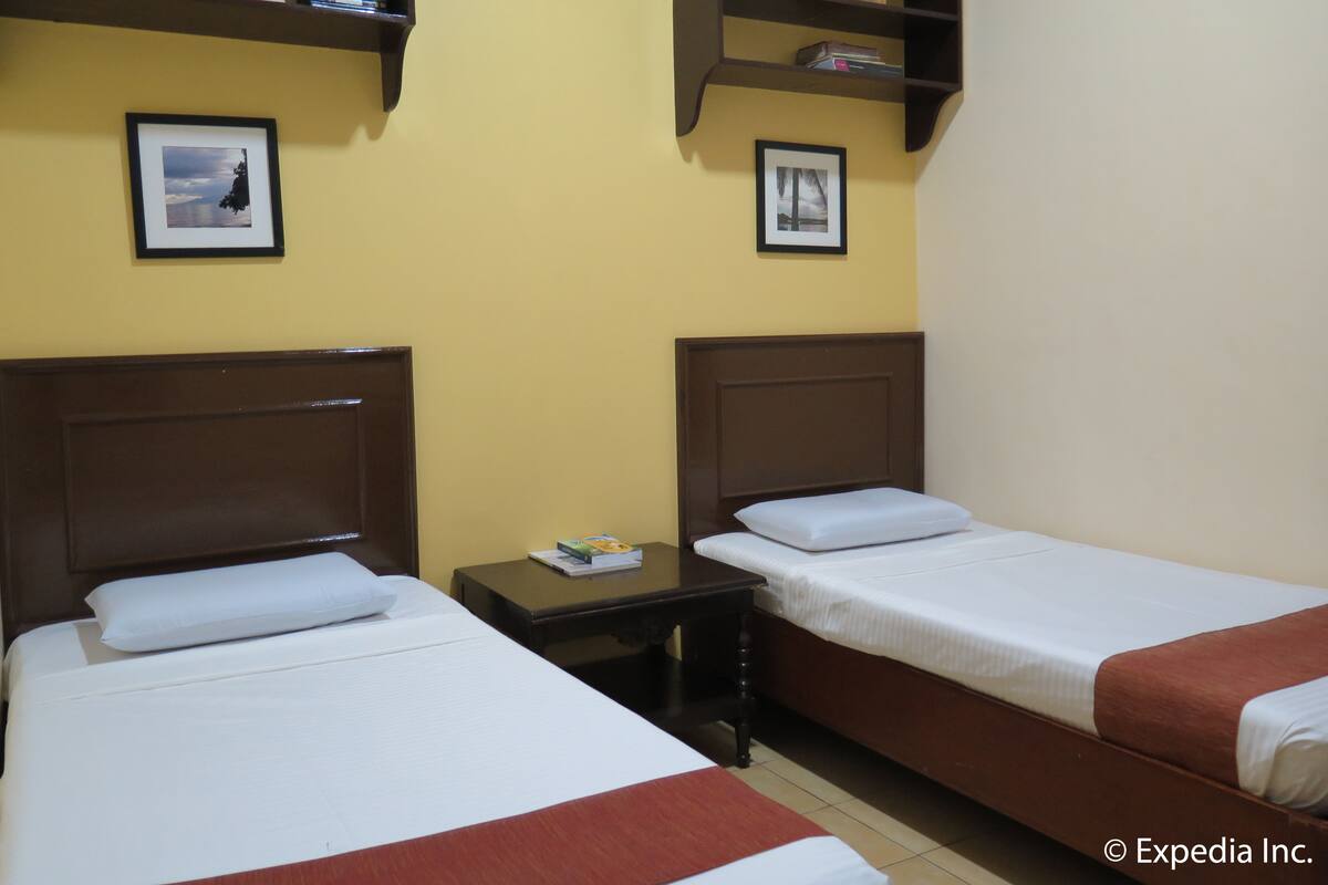 Quarto de hotel com duas camas de solteiro, uma mesa de cabeceira, uma pequena mesa e duas prateleiras fixadas na parede com livros e quadros.