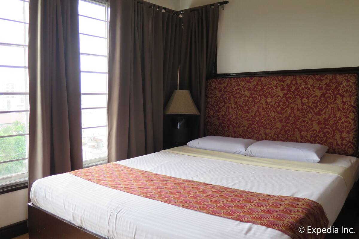 Quarto de hotel com uma cama grande, cabeceira estampada, janela com cortinas e mesa de cabeceira com abajur.