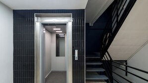Um elevador moderno com acabamento metálico, uma escadaria com guarda-corpos pretos e uma parede revestida de azulejos.