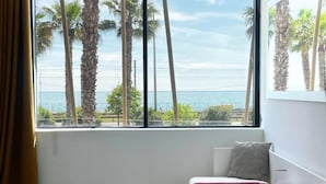 Quarto com uma cama, janela com vista para palmeiras e o mar, e uma pequena mesa de cabeceira.