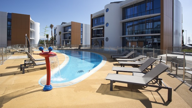 Piscina com espreguiçadeiras, uma fonte de água e edifícios modernos com janelas amplas.