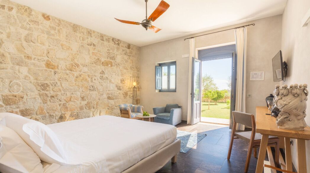 Quarto com cama, ventoinha no teto, parede de pedra, janela e vista para o exterior.