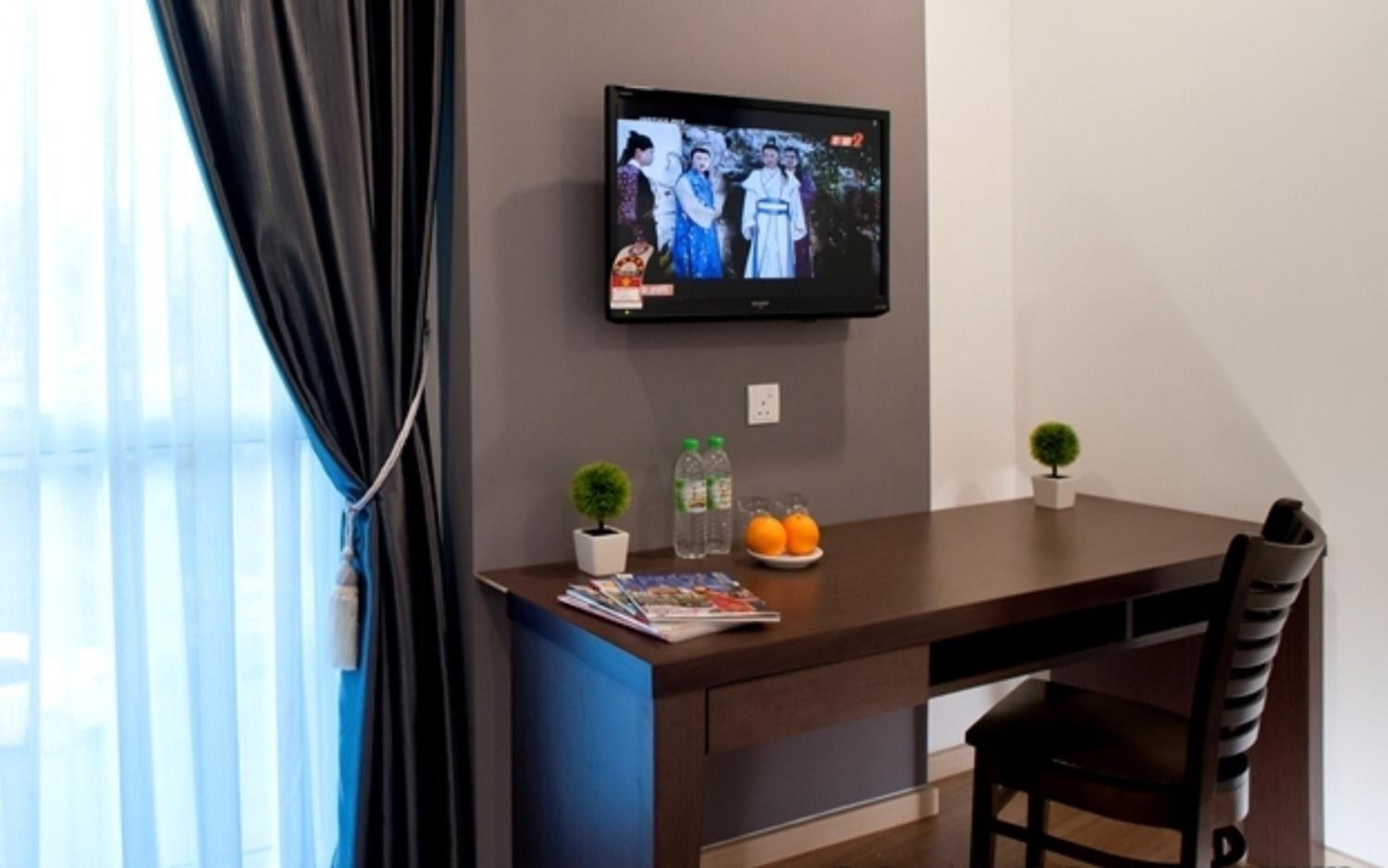 Quarto de hotel com televisão na parede, uma mesinha com uma revista, duas plantas em vasos e um prato com laranjas.