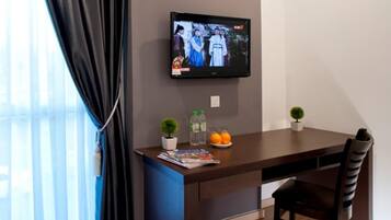 Quarto de hotel com televisão na parede, uma mesinha com uma revista, duas plantas em vasos e um prato com laranjas.