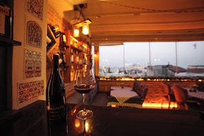 Uma área de bar com uma garrafa de vinho e um copo, vista da cidade através de amplas janelas, e um espaço de estar aconchegante com almofadas.