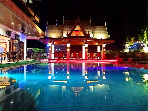 Vista noturna de uma área de piscina de hotel com um pavilhão tradicional, assentos e um bar.