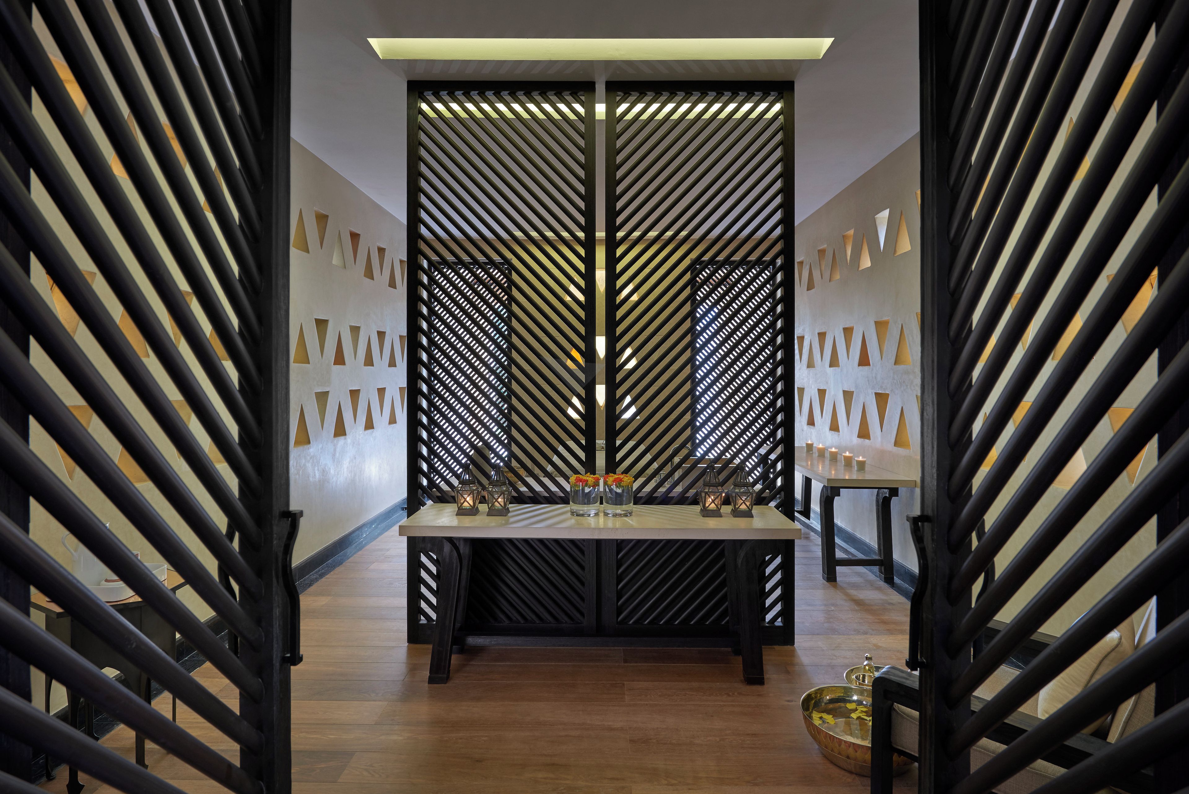 Um quarto com uma mesa central, um separador decorativo com padrões geométricos e uma série de recortes triangulares na parede.