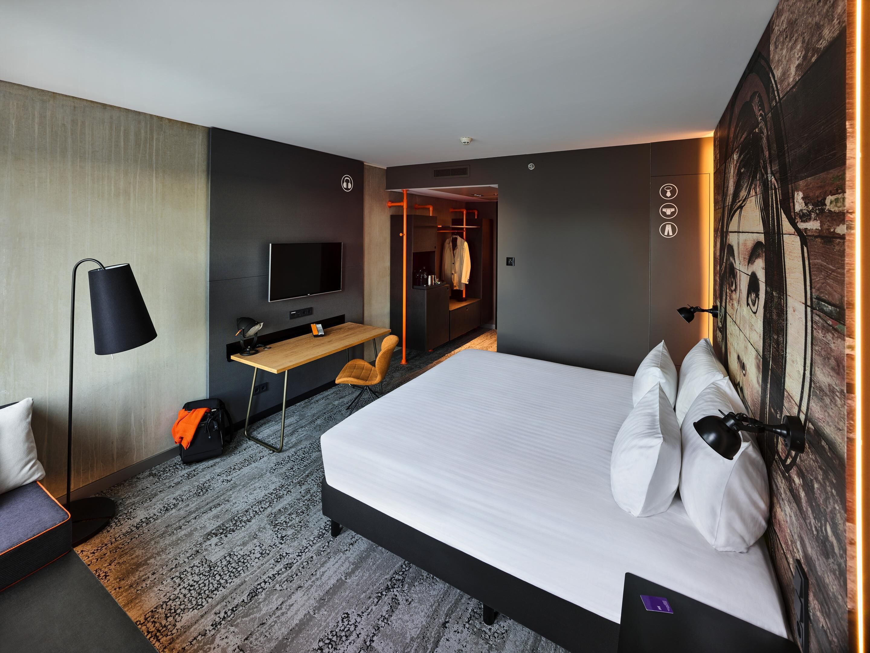 Quarto de hotel moderno com uma cama grande, uma escrivaninha com televisão, um guarda-roupa e um quadro na parede.