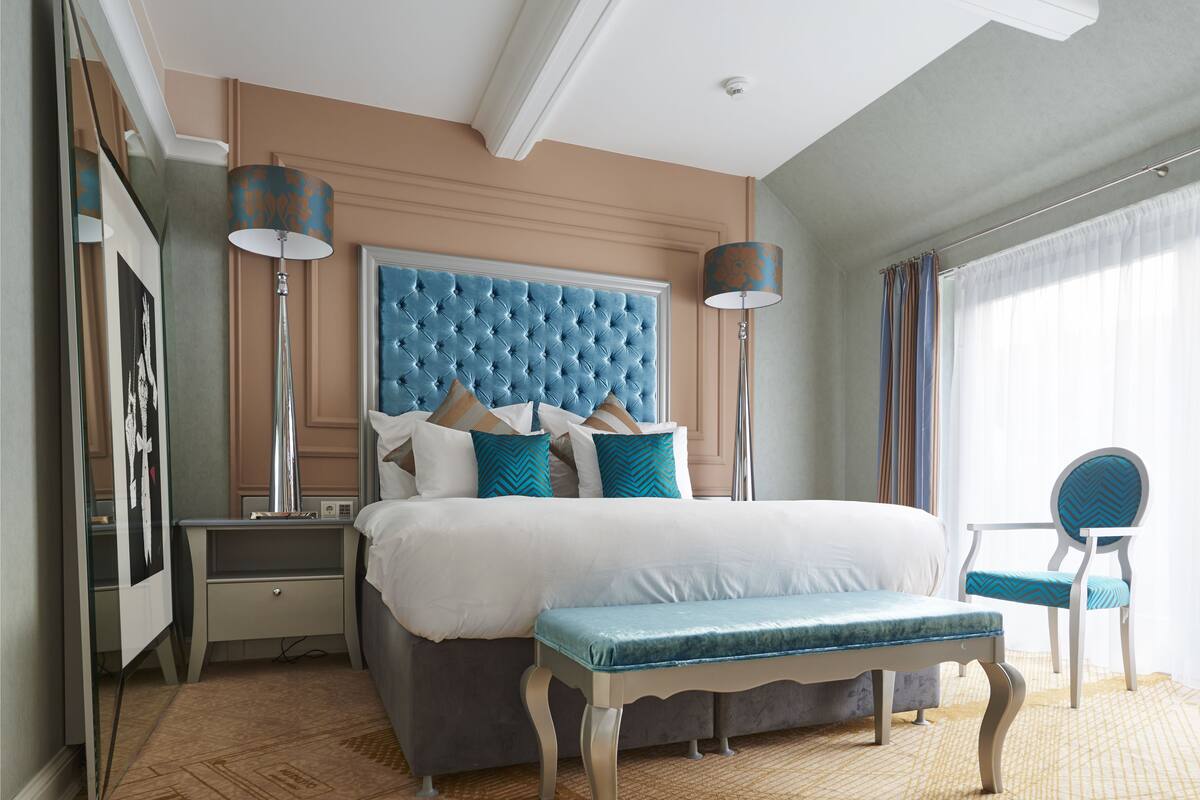 Quarto de hotel moderno com uma cama grande, um banquinho, uma mesa de cabeceira, um abajur e uma janela ampla.