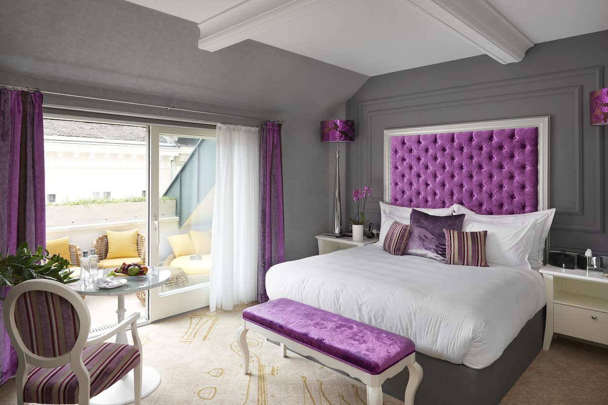Quarto moderno com uma cama grande, um banco, área de refeições com mesa e cadeiras, e vista pela janela.