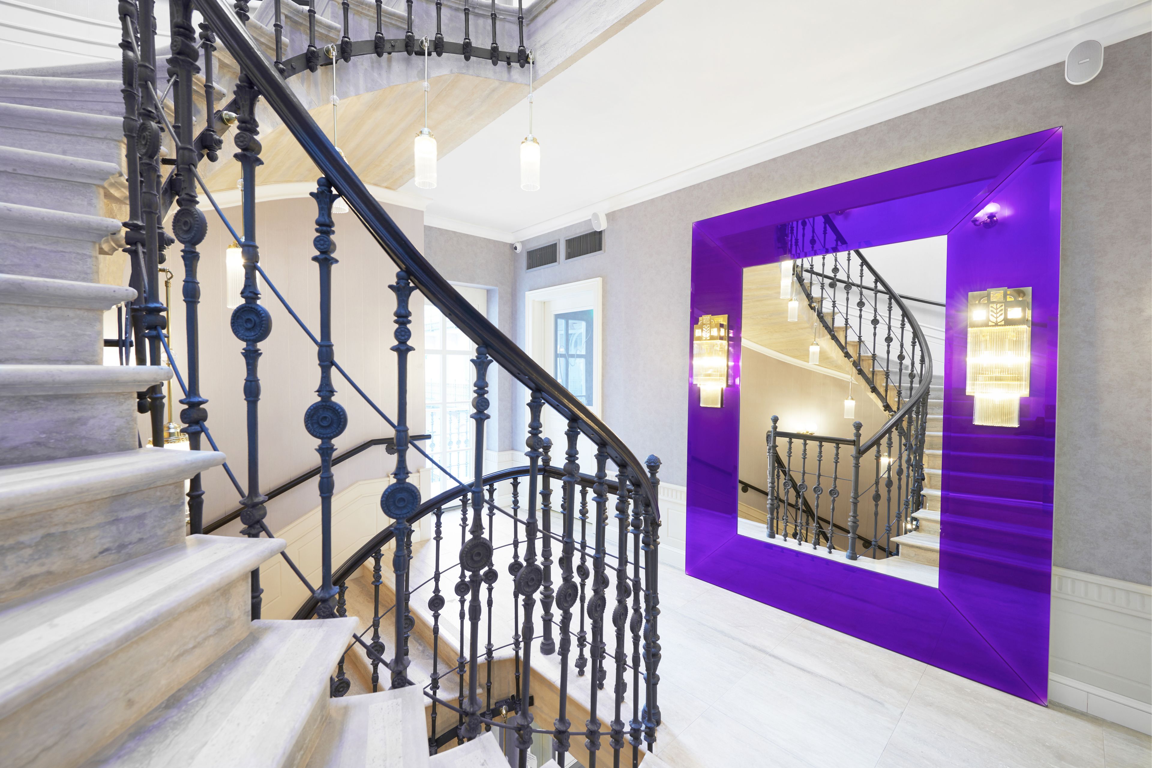 Uma escadaria imponente com balaústres pretos ornamentados e degraus de mármore, que leva a um patamar com parede em tons de roxo e uma parte espelhada.
