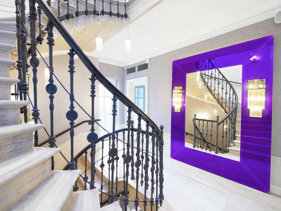 Uma escadaria imponente com balaústres pretos ornamentados e degraus de mármore, que leva a um patamar com parede em tons de roxo e uma parte espelhada.