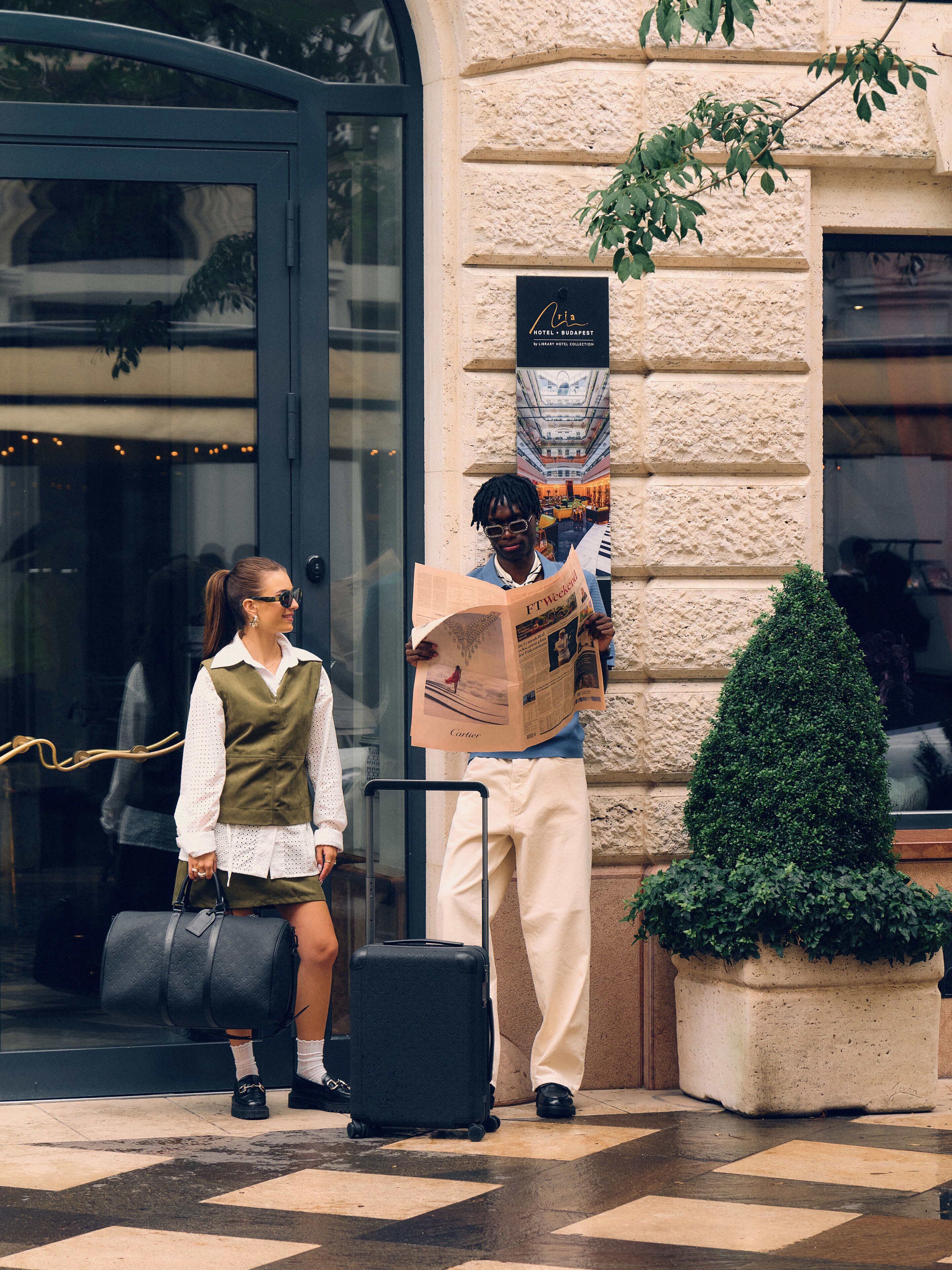 Um homem carregando uma caixa grande e uma mala, parado ao lado da entrada de um edifício com uma placa que diz "Atria Hotel Budapest".