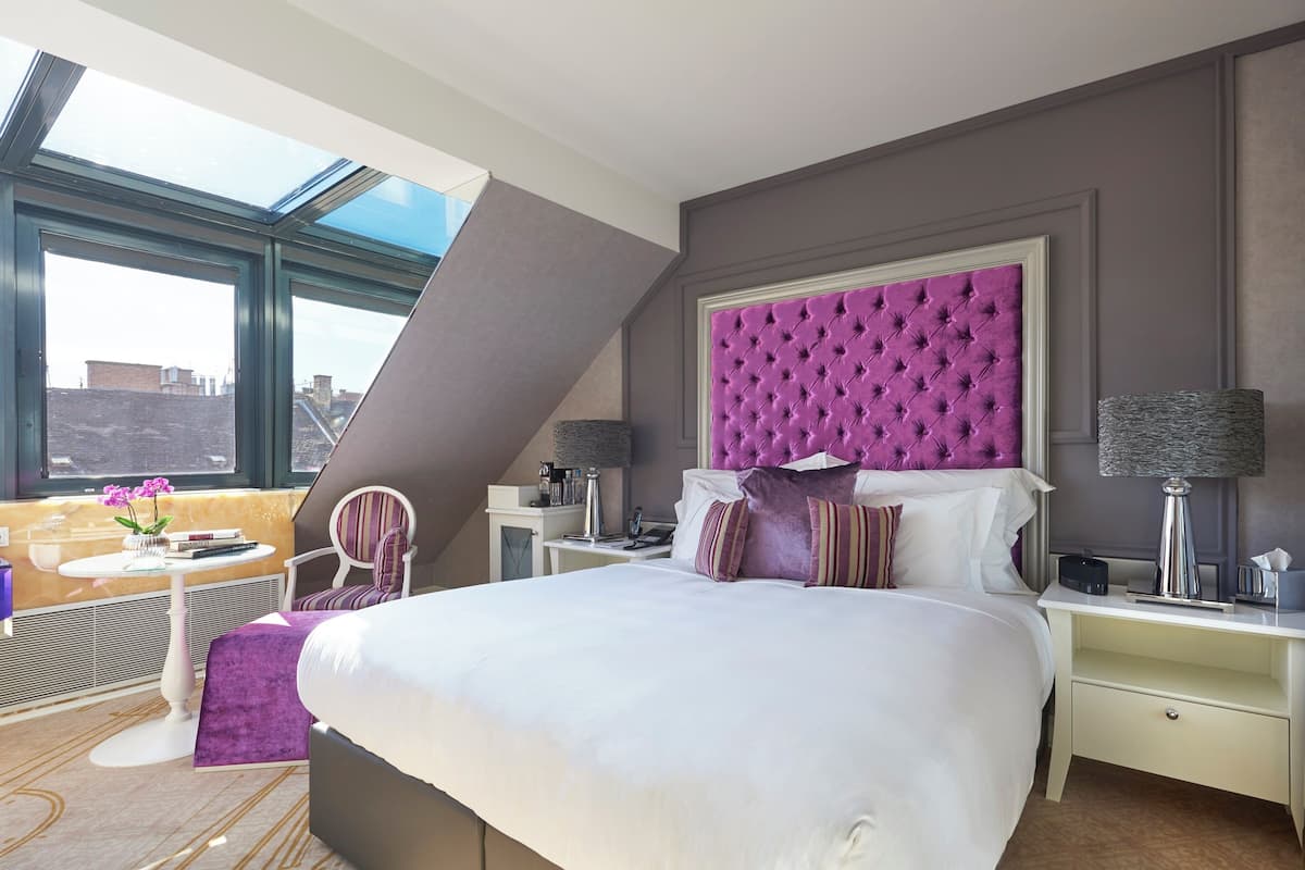 Quarto de hotel moderno com uma cama grande, uma escrivaninha, uma cadeira e vista para a cidade através de janelas amplas.