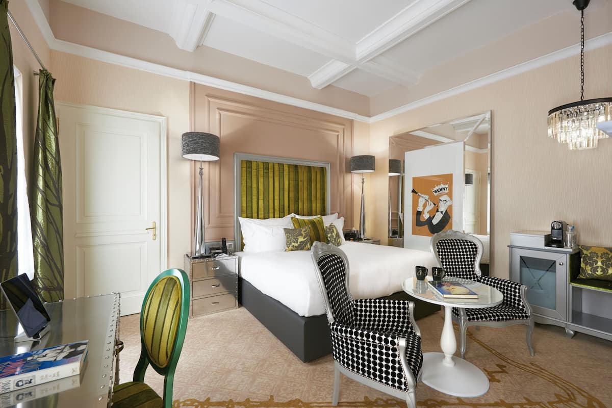Quarto de hotel com cama, duas cadeiras, uma mesa pequena, escrivaninha com abajur, espelho e lustre.
