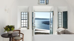 Quarto com cama, poltrona, mesa com planta e duas janelas com vista para o mar.