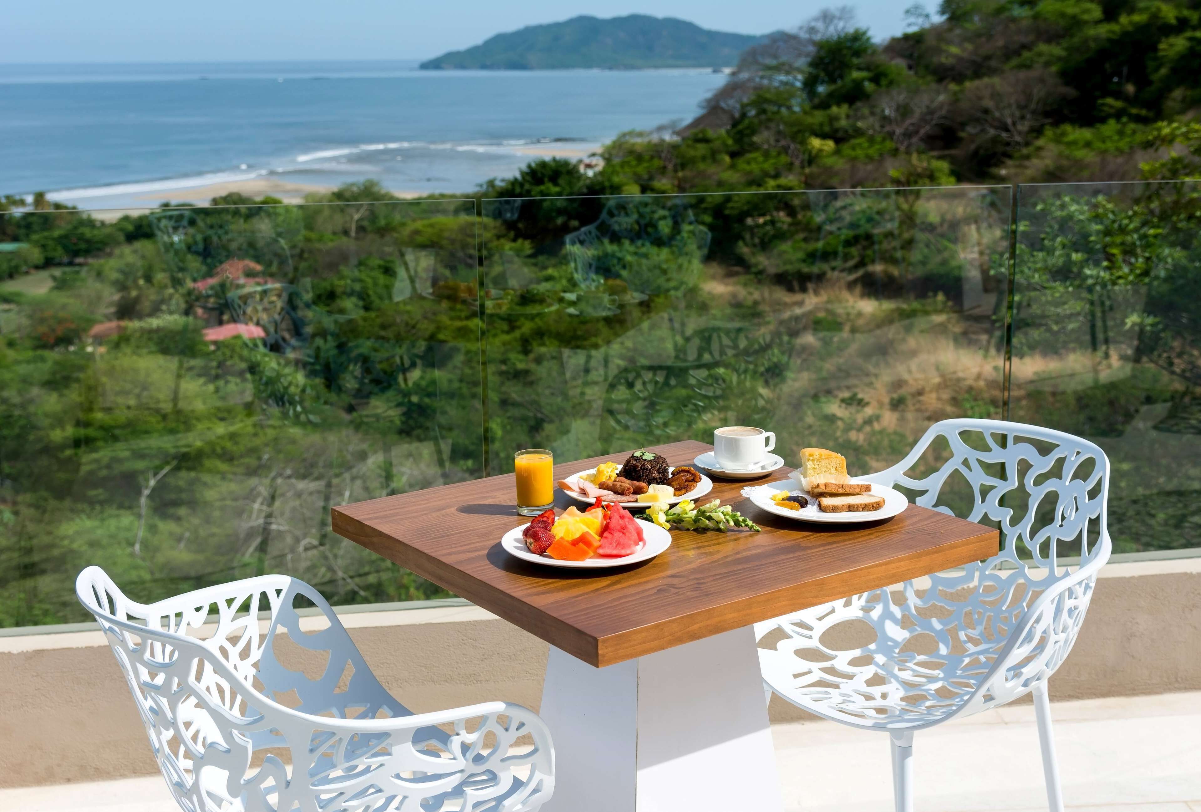 Mesa com itens de café da manhã e um copo de suco de laranja numa varanda com vista para uma praia e área arborizada.