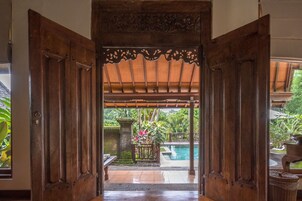 Uma porta de madeira dá acesso a uma área externa coberta, com piscina e vegetação exuberante.