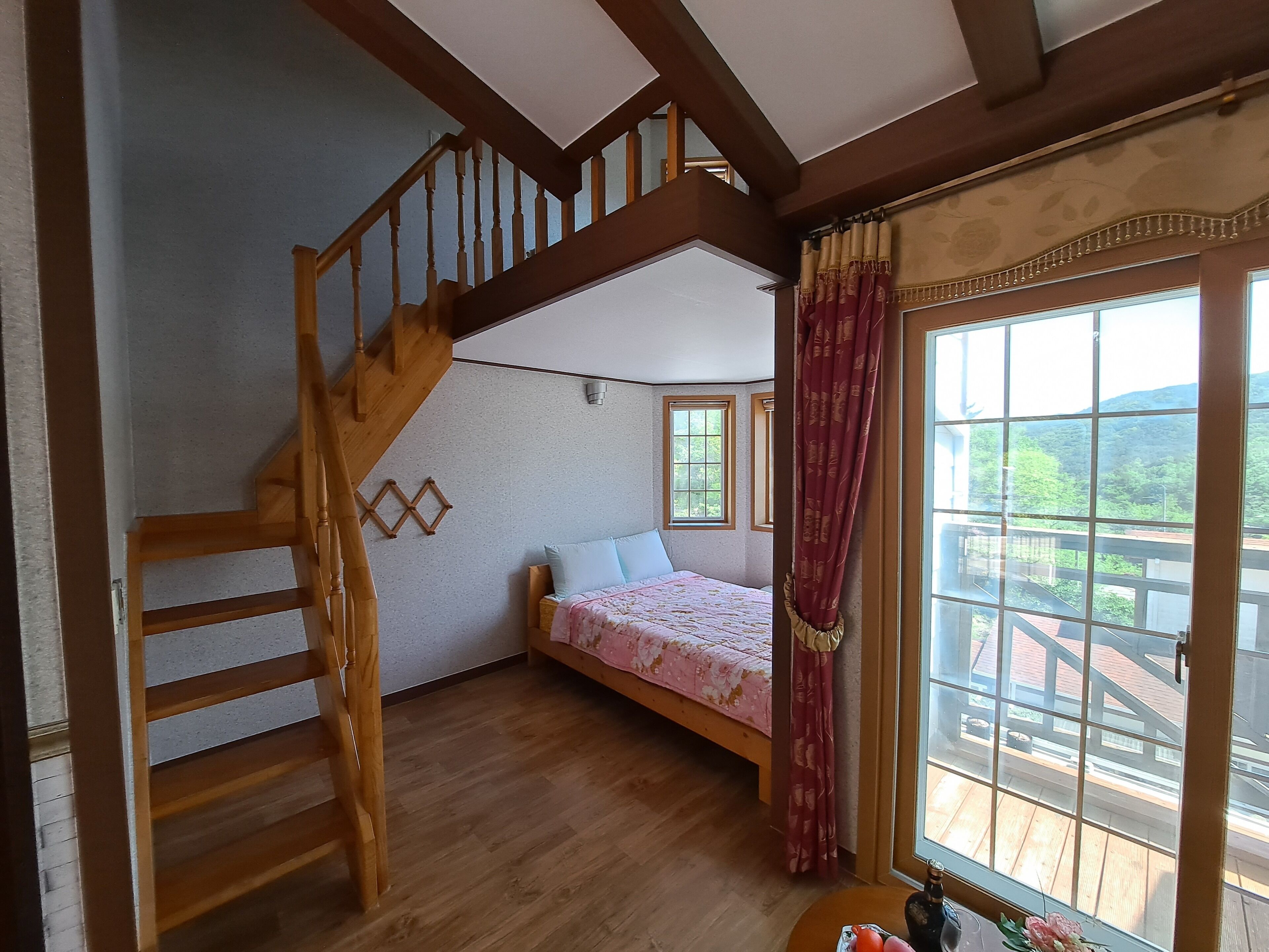 Um quarto com escada de madeira, cama com colcha florida, janela com vista para um deck de madeira e luminária fixada na parede.