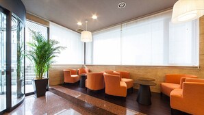 Um lobby com assentos laranja, uma grande planta em vaso e vista da cidade através de grandes janelas.