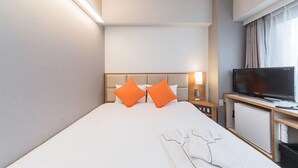 Quarto de hotel com cama, duas almofadas laranja, mesa de cabeceira com abajur, televisão e pequeno frigorífico.