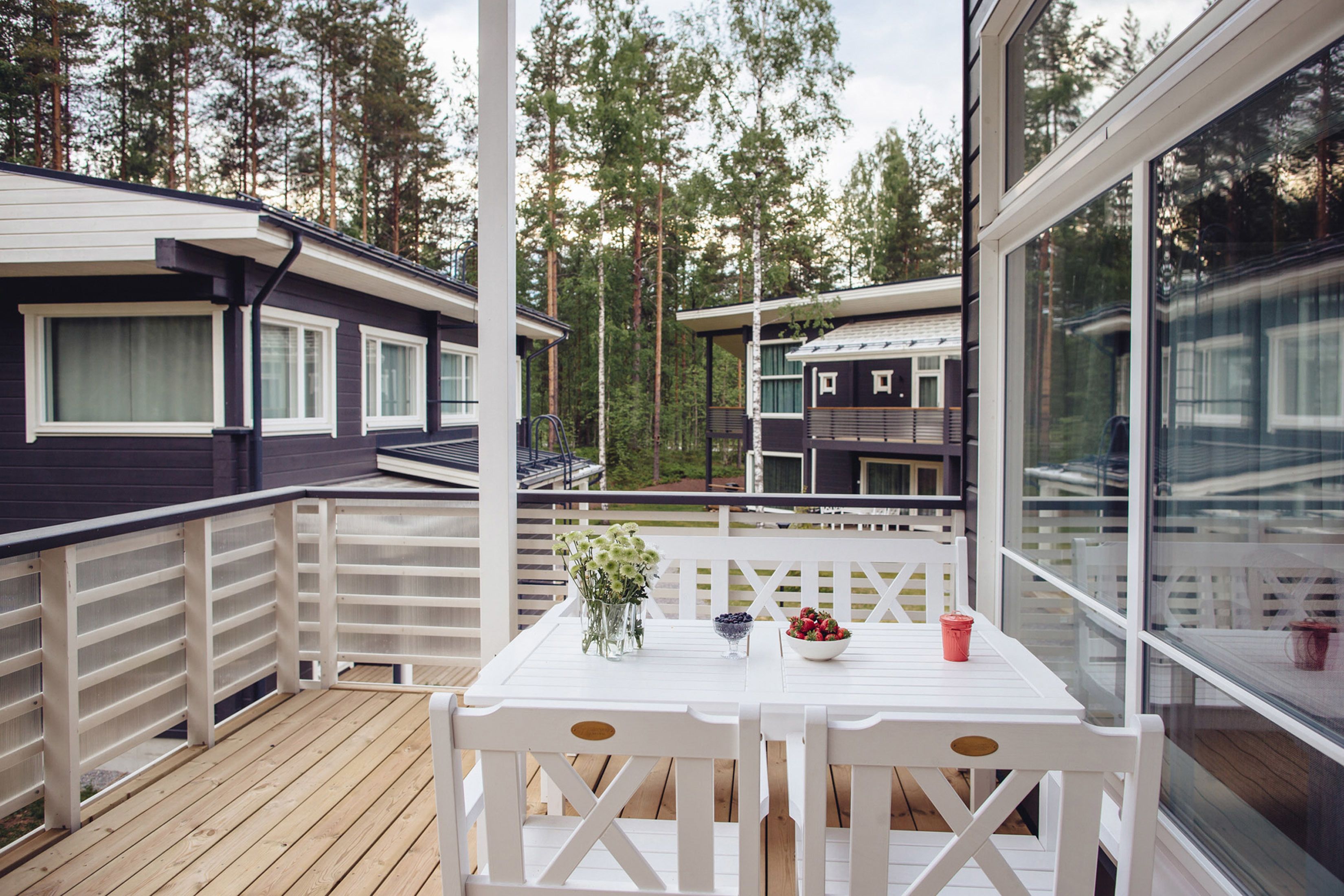 Um terraço com mesa e cadeiras brancas, cercado por um corrimão branco, com vista para uma área arborizada.